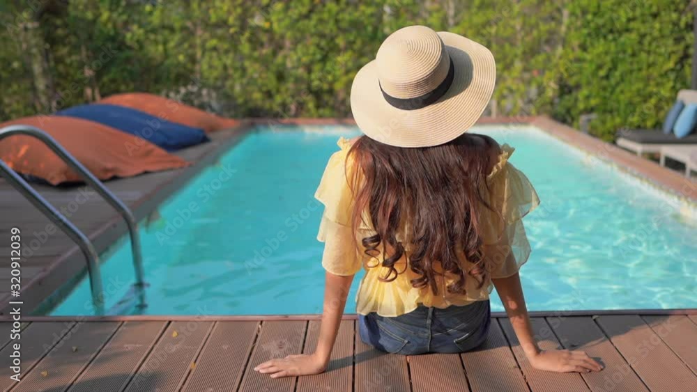 Back view of a female tourist sitting at a swimming pool wearing a sun ...