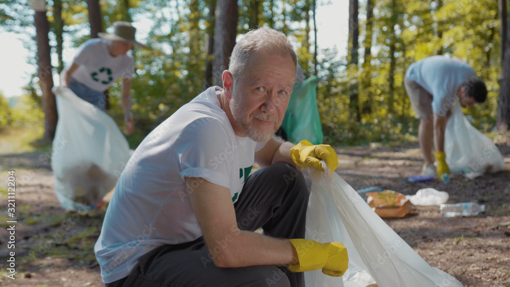 Portrait senior man cleans forest of debri folding it in plastic bag ...