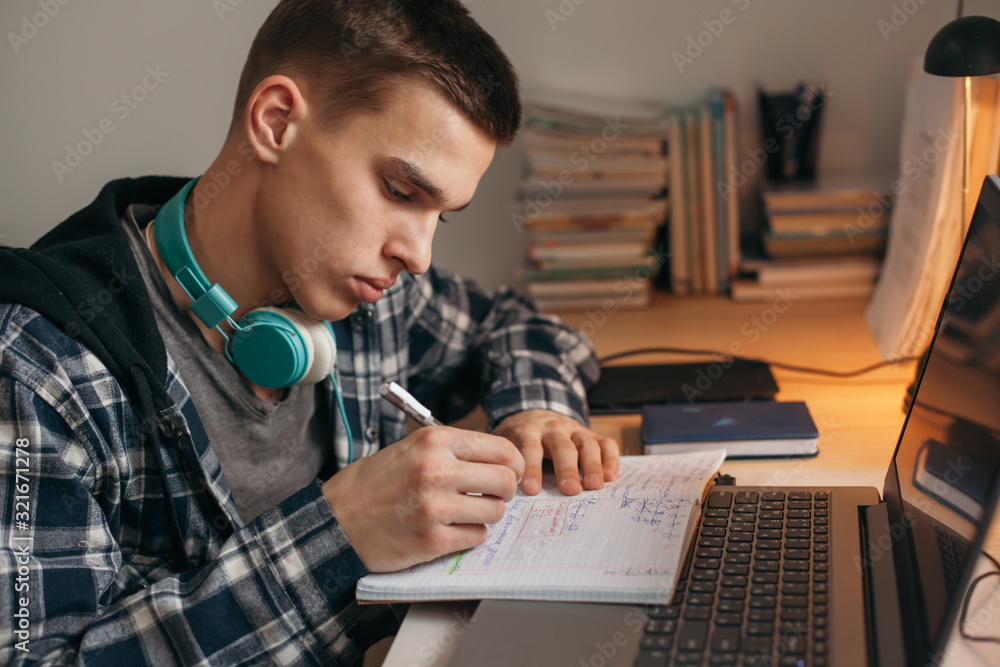 Teenage boy doing homework using computer sitting by desk in room alone ...
