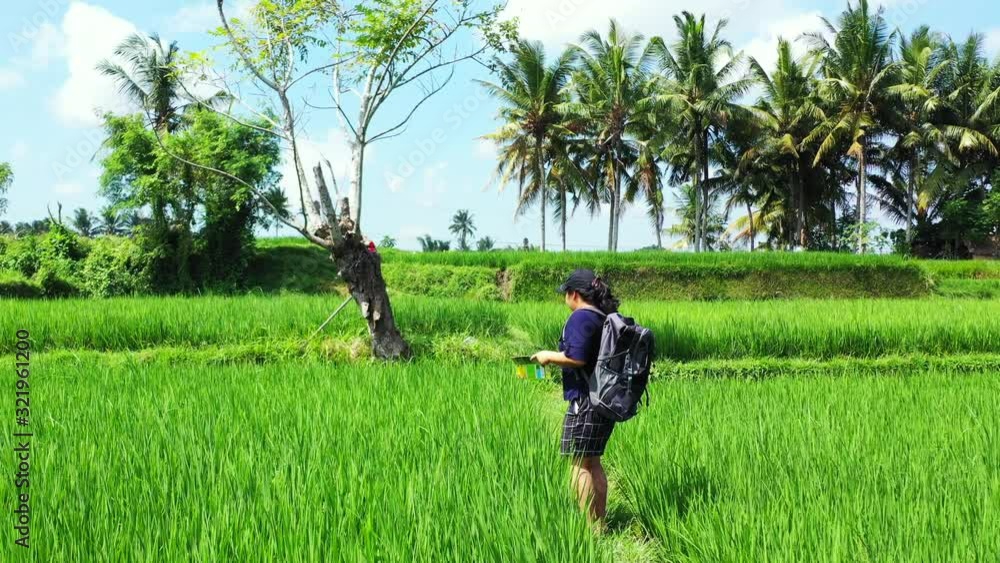 A young woman in a lush green paddy field with backpack and map ...