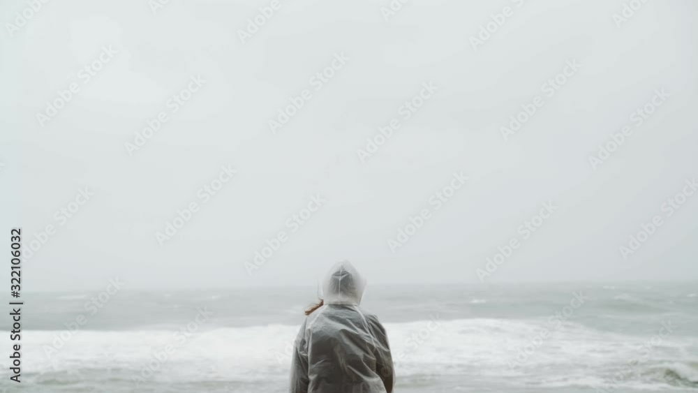 A girl in a white raincoat is walking along the stormy sea. A woman is ...