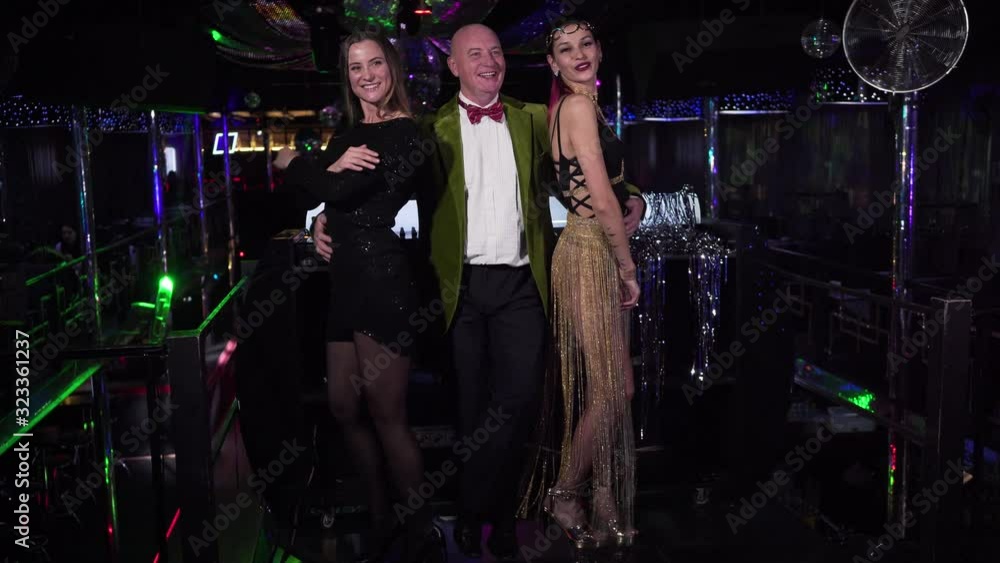 Elderly Caucasian man and two young women posing in night club ...