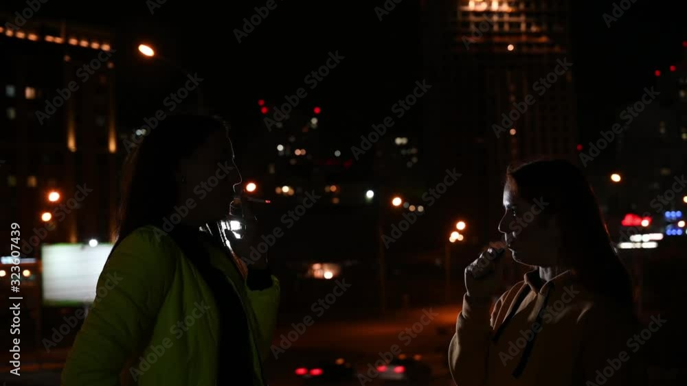 Two girlfriends smoke on the background of the night city. Women smoke ...