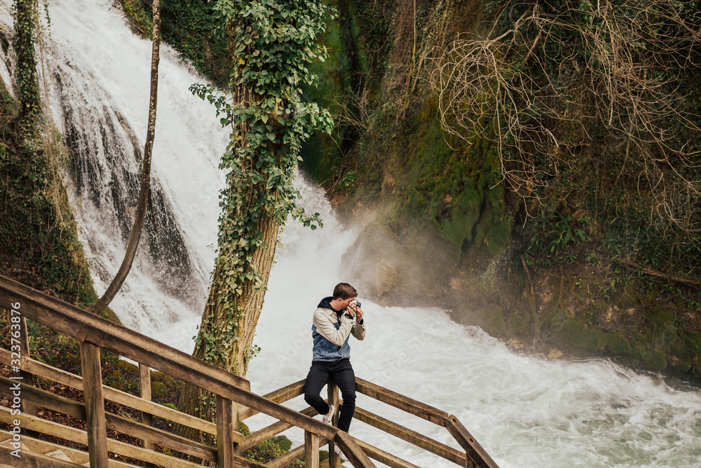 Young stylish tourist are photographing waterfall. Man sitting in front ...