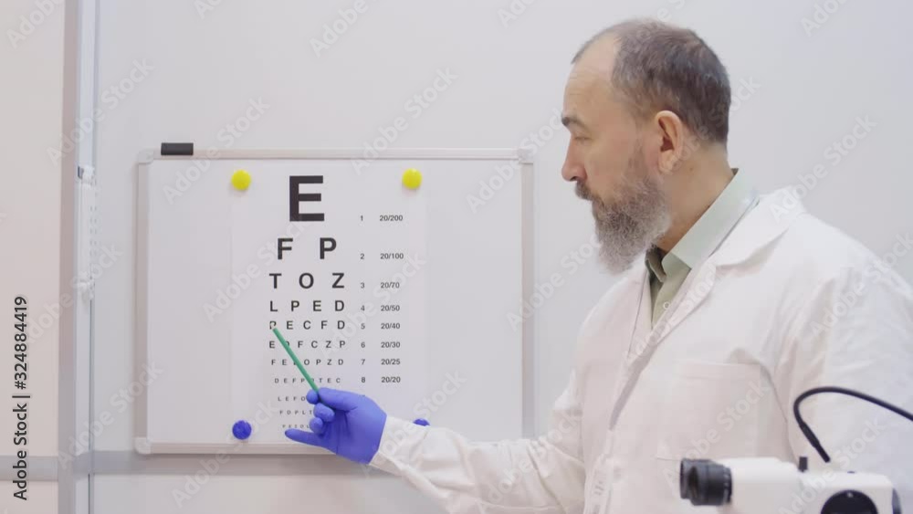 Professional elderly optometrist in lab coat pointing at eyechart with ...