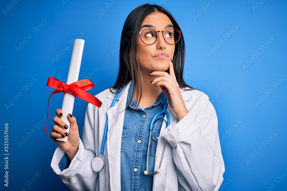 Young beautiful brunette doctor woman wearing glasses and coat holding ...