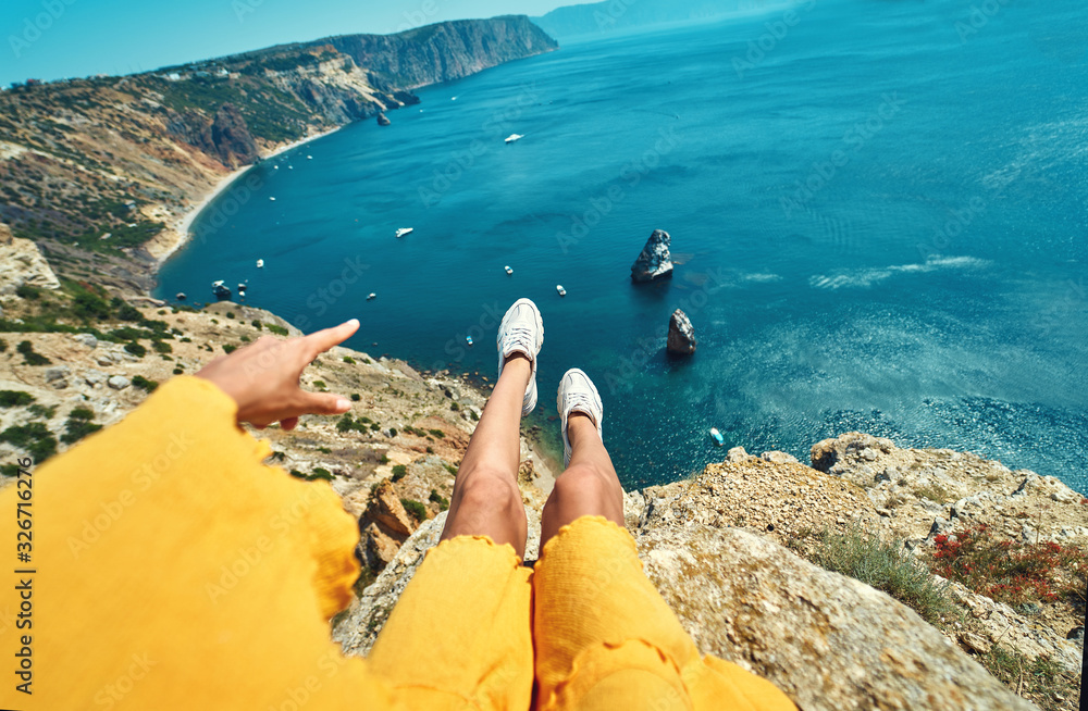 first person view of taned woman in yellow dress sitting on on cliff ...