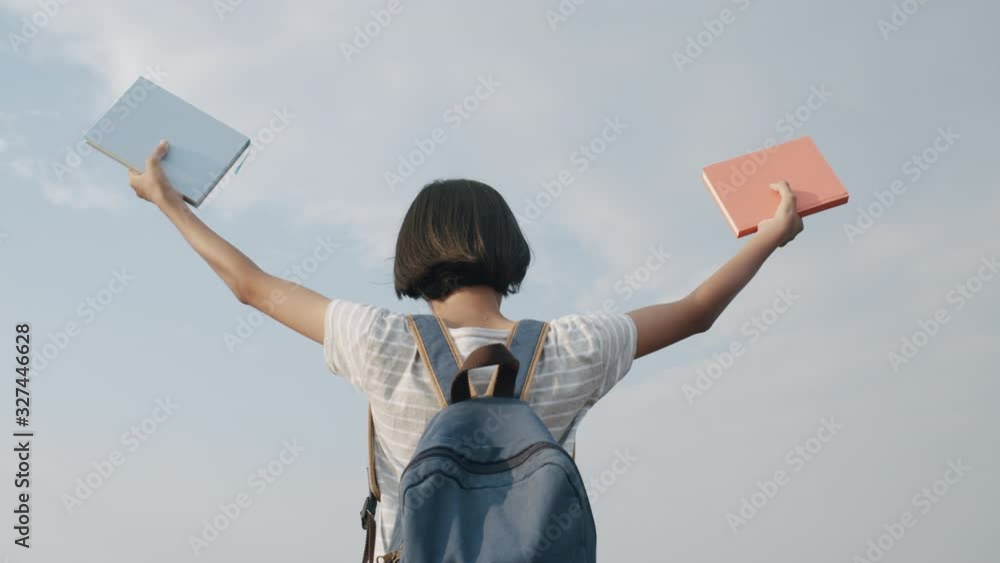 Back view of Asian girl holding a book and hand raised up to blue sky ...