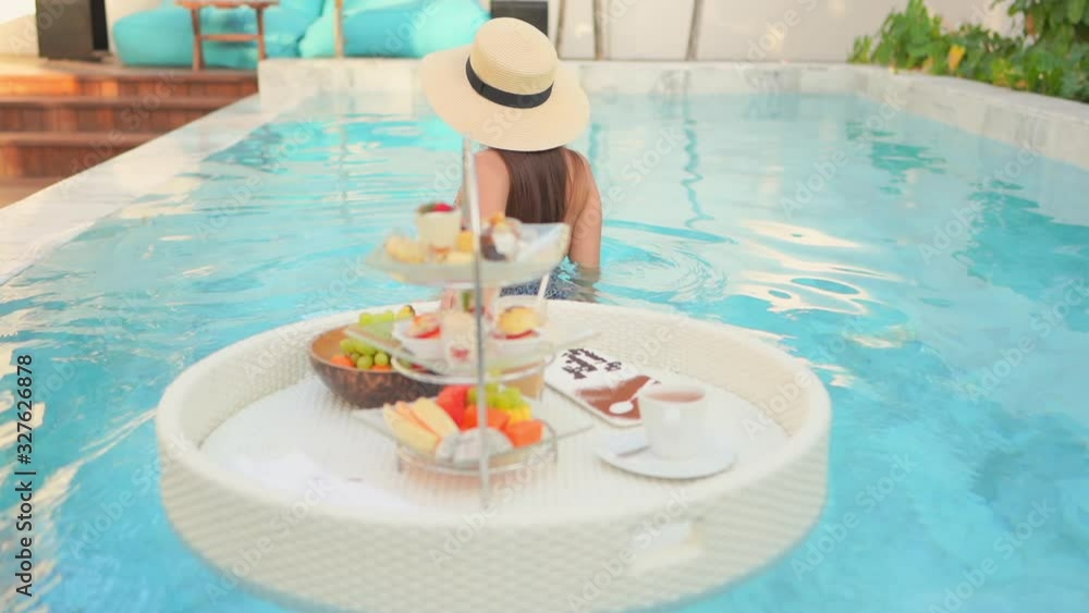 A young Asian woman in a resort swimming pool guides a floating platter ...