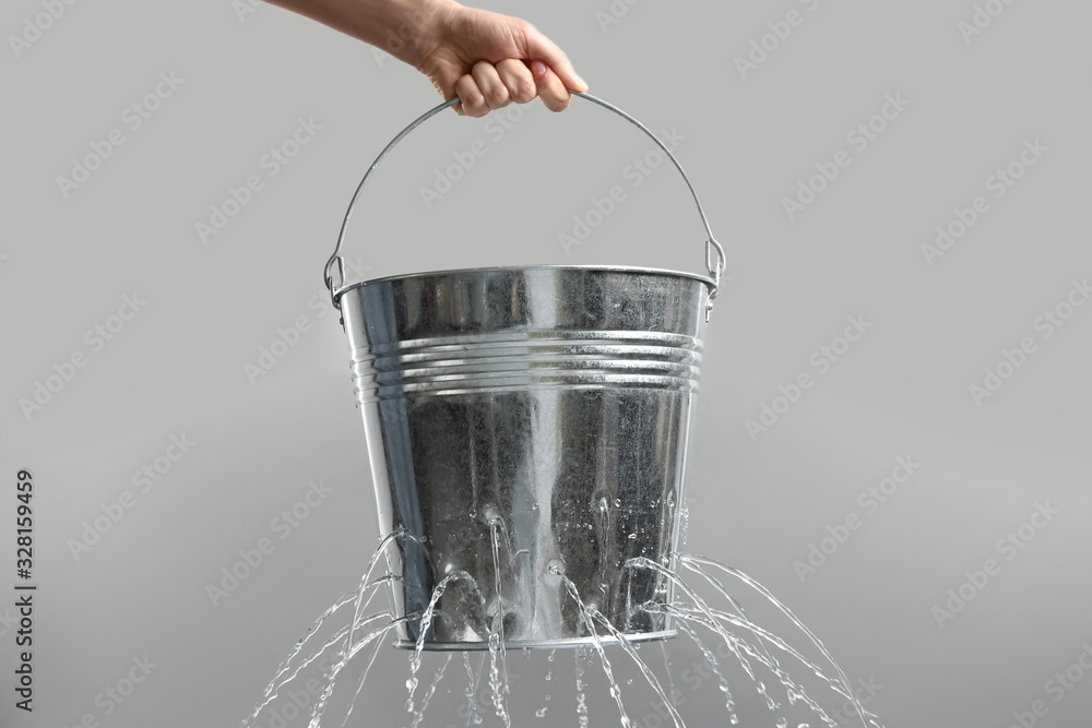 Woman holding leaky bucket with water on light grey background, closeup ...