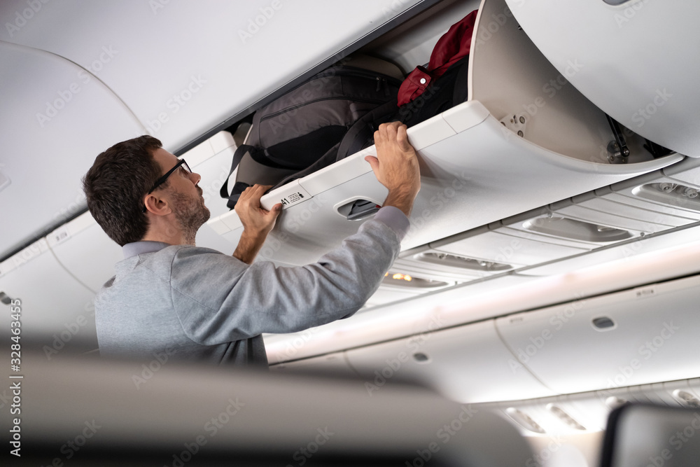 Young man putting luggage into overhead locker on airplane. Traveler ...