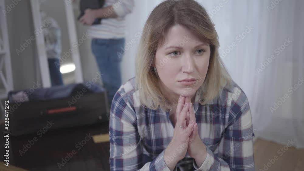 Upset young Caucasian woman sitting on bed holding hands clasped as unrecognizable man packing ...