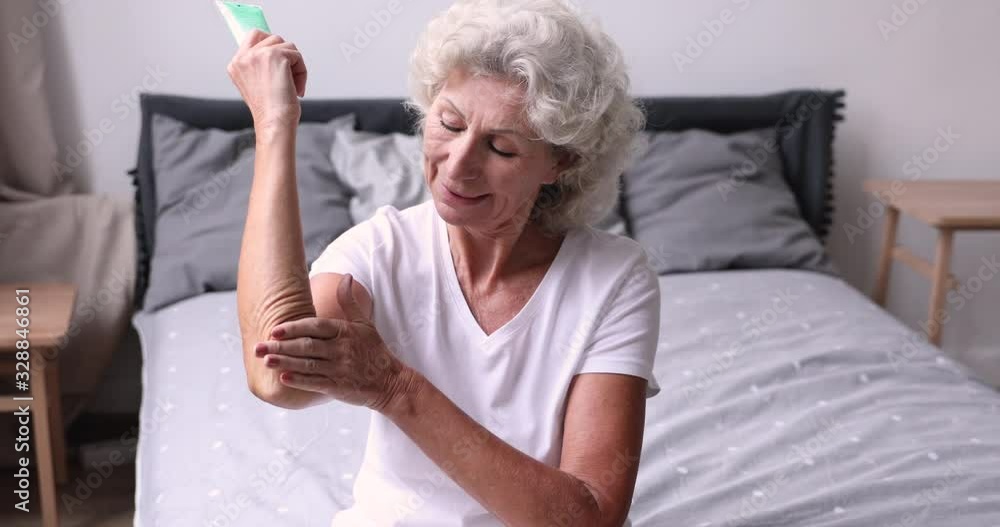 Smiling older woman applying moisturizer cream on elbow sitting on bed ...