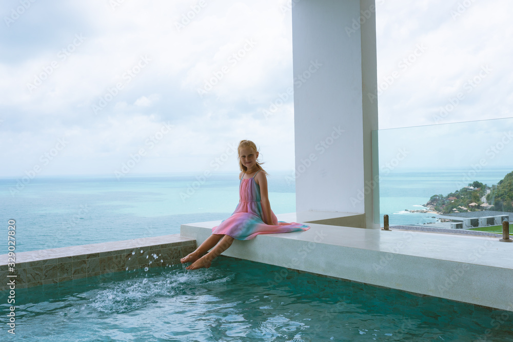 Little and beautiful girl with red hair and freckles sits by the pool ...