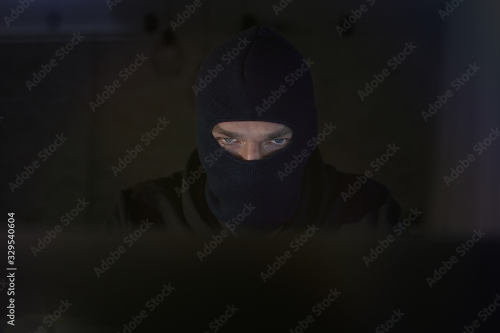 Male hacker in the hood in a dark room using computer virus program for cyber attack. Hackers ...