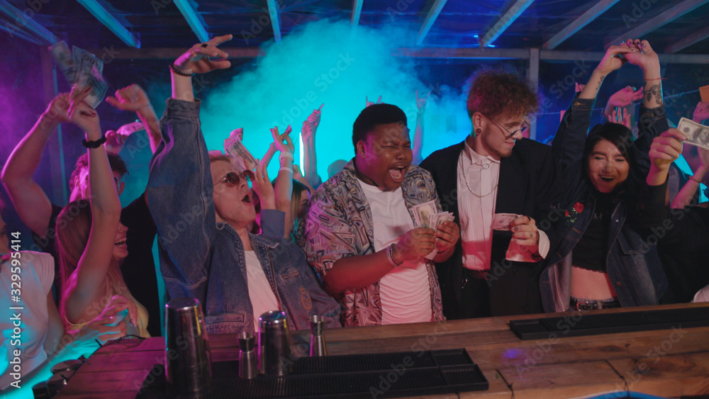 Attractive and joyful young friends holding money up dancing off at bar ...