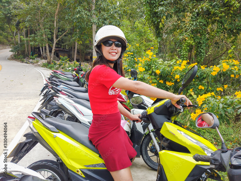 Young Thai lady wearing an helmet. Behind her, a green motorcycle and a tropical background ...