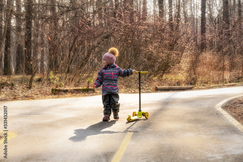 child with a scooter on an asphalt road in a spring park in a hat with ...