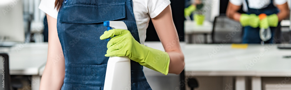 cropped view of young cleaner in overalls holding spray bottle with ...