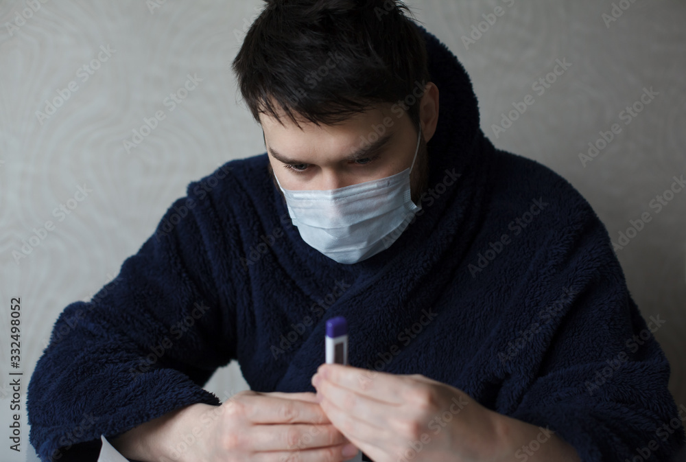Sad man checking his temperature. Man in a medicine mask and home gown ...