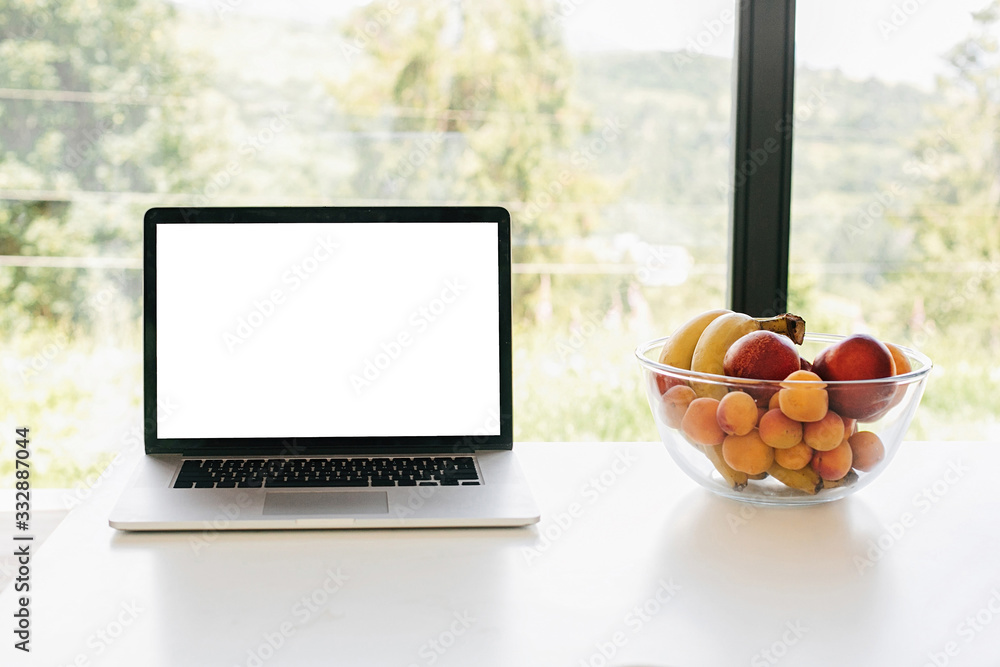 Laptop with blank white screen and fruits in glass bowl on countertop ...