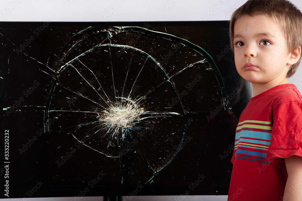 Sad little boy standing in front of a TV with broken screen after an ...