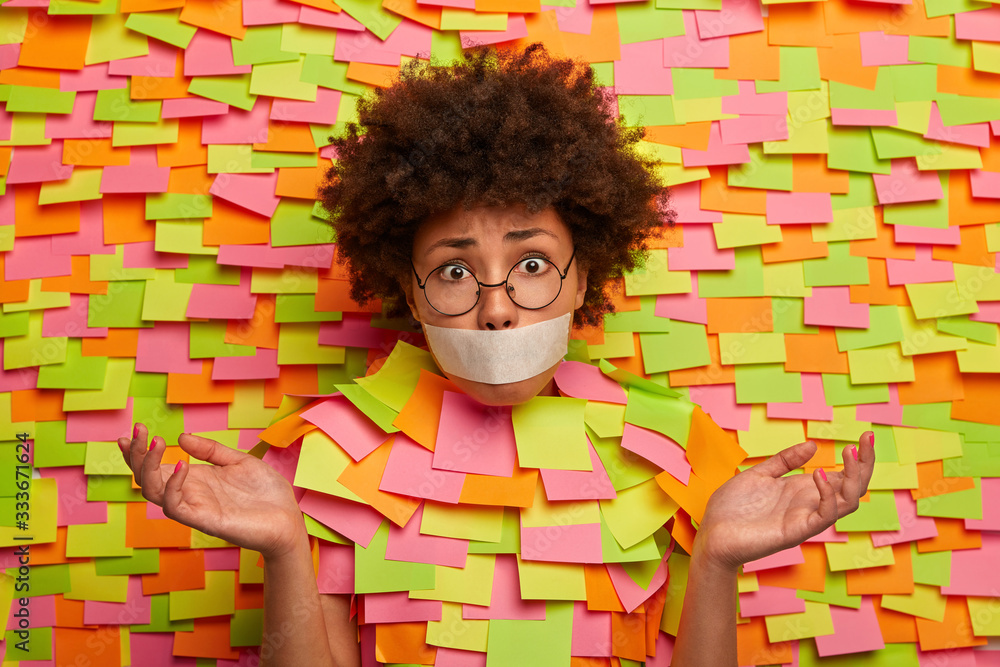 Confused clueless Afro American woman with taped mouth spreads palms ...