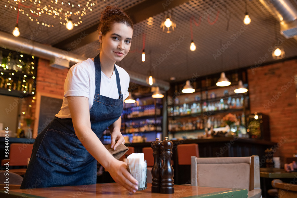 Pretty young waitress bending over table while putting glass with ...
