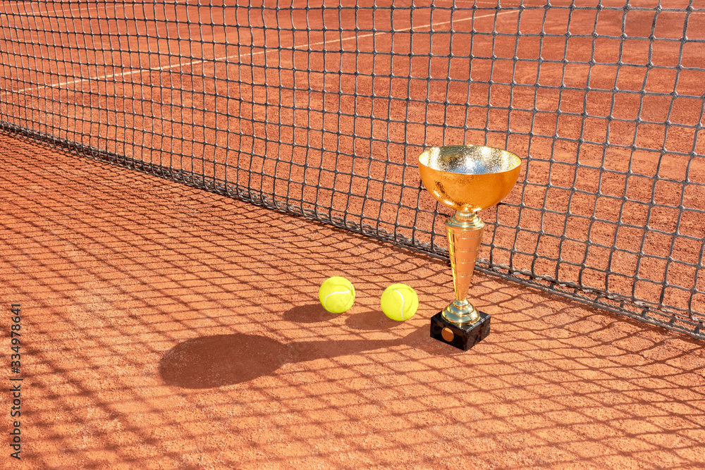 Trophy cup and tennis balls on a red clay court and shadow from the net ...