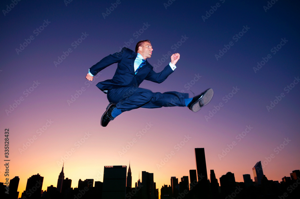 Businessman superhero leaping outdoors at dusk above a dusk city ...