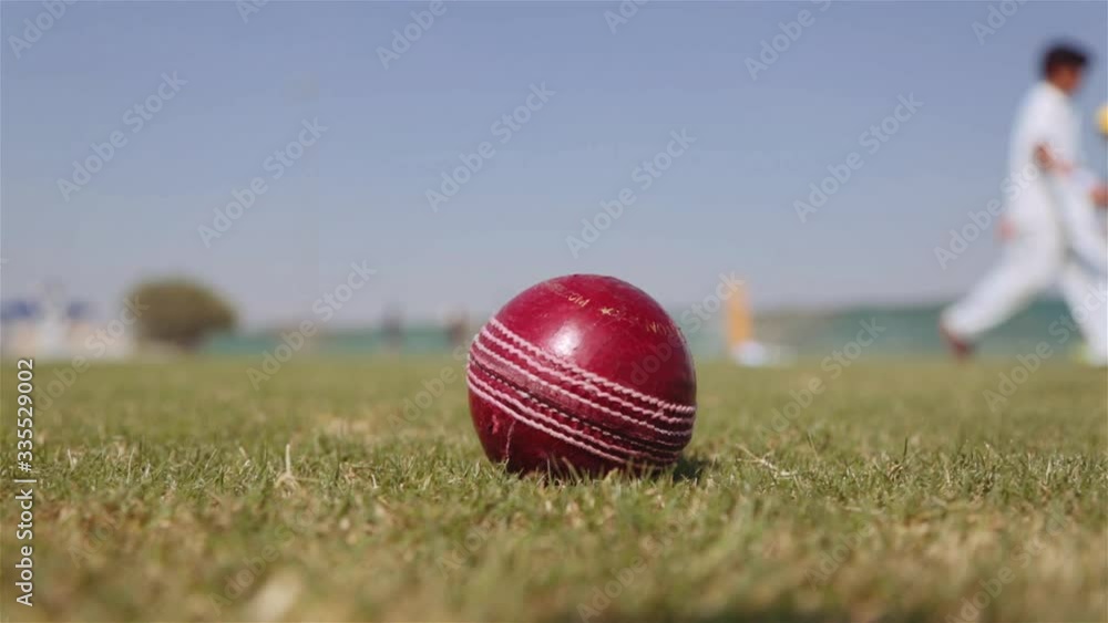 Red cricket ball roll into frame with people playing cricket in ...