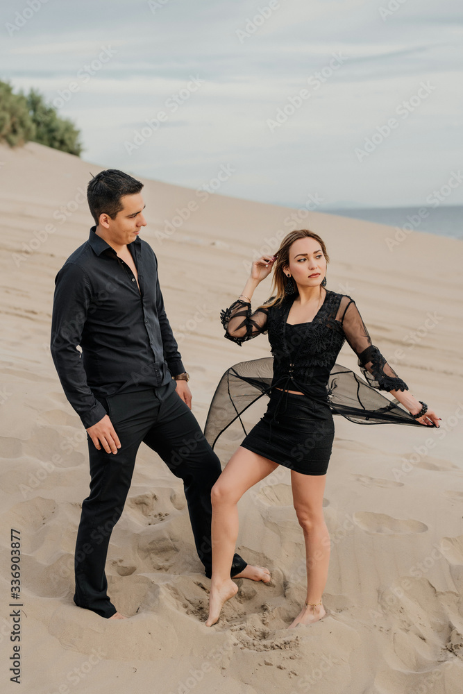 Pareja modelos enamorados vestidos de negro, parados sobre la arena con ...