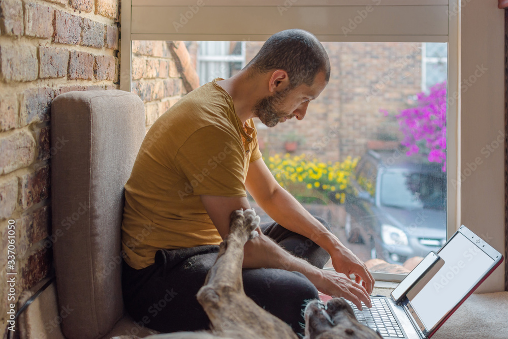 Young man in sweat pants and t-shirt typing in his laptop while his dog ...