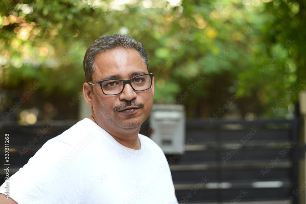 Indian man wearing spectacles in a white t-shirt outside a gate against ...