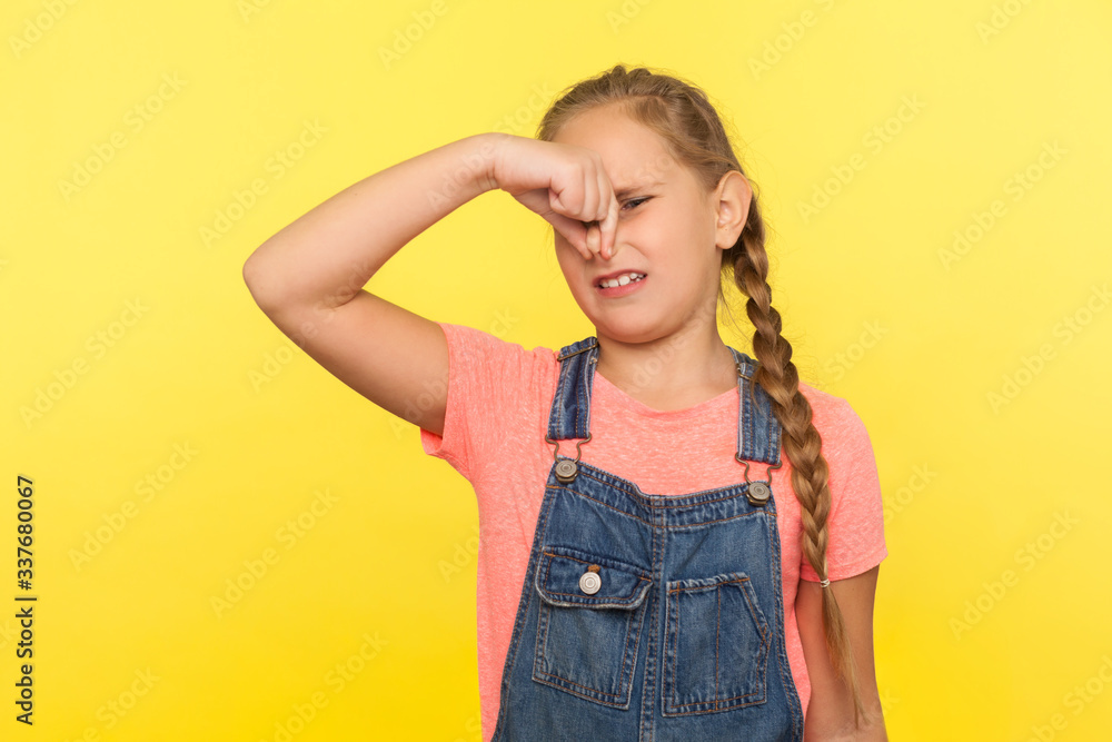 Awful smell. Portrait of little girl with braid in denim overalls ...