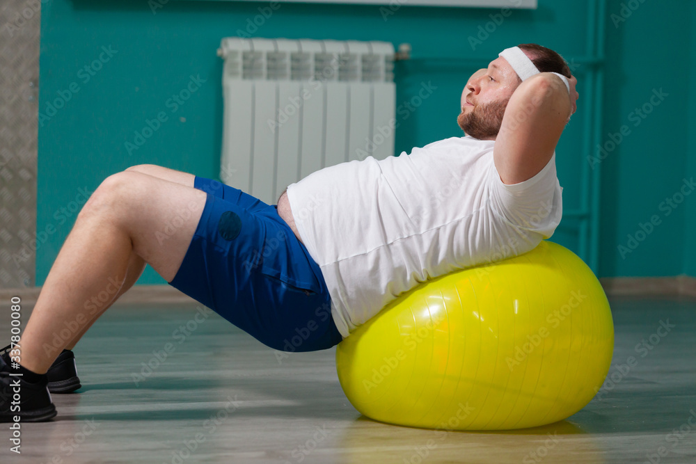 Overweight man is lying on a fitness ball during group fitness classes ...
