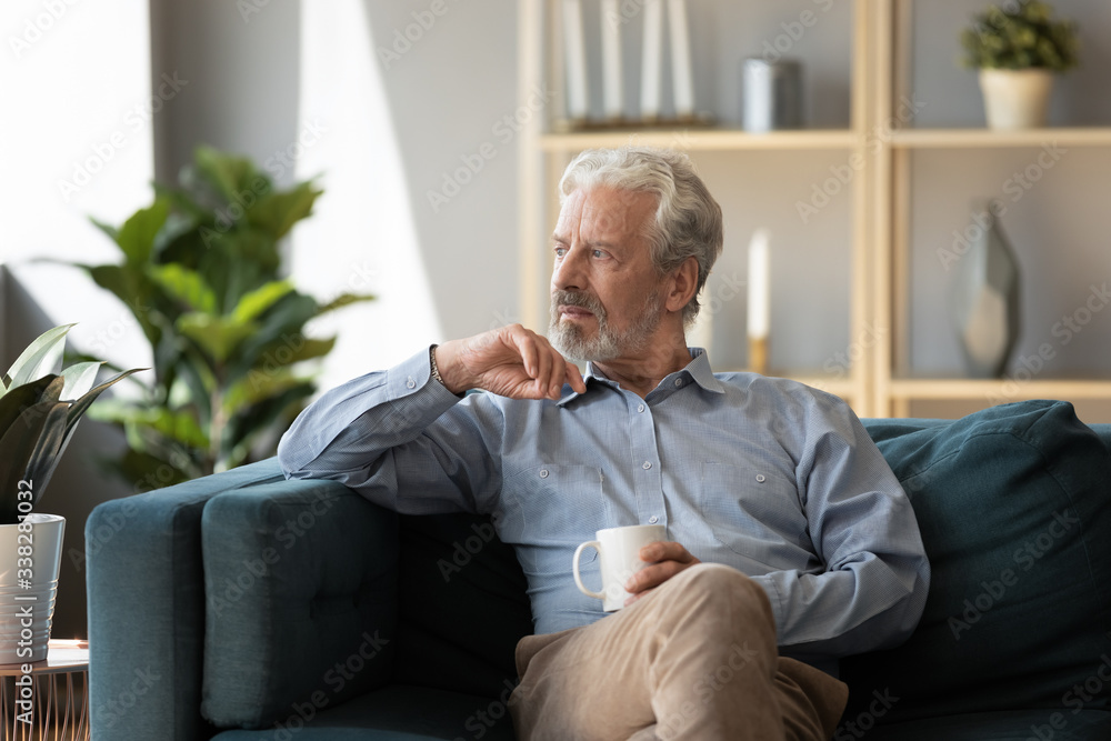 Thoughtful older man sitting on couch at home alone, holding cup of tea ...