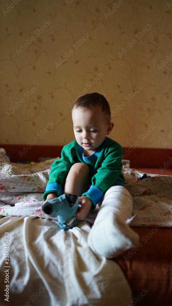 A small cute child with plaster cast on leg sitting on untidy bed at ...
