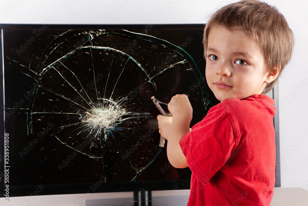 Cute little boy standing in front of a TV with broken screen and aiming with his slingshot ...