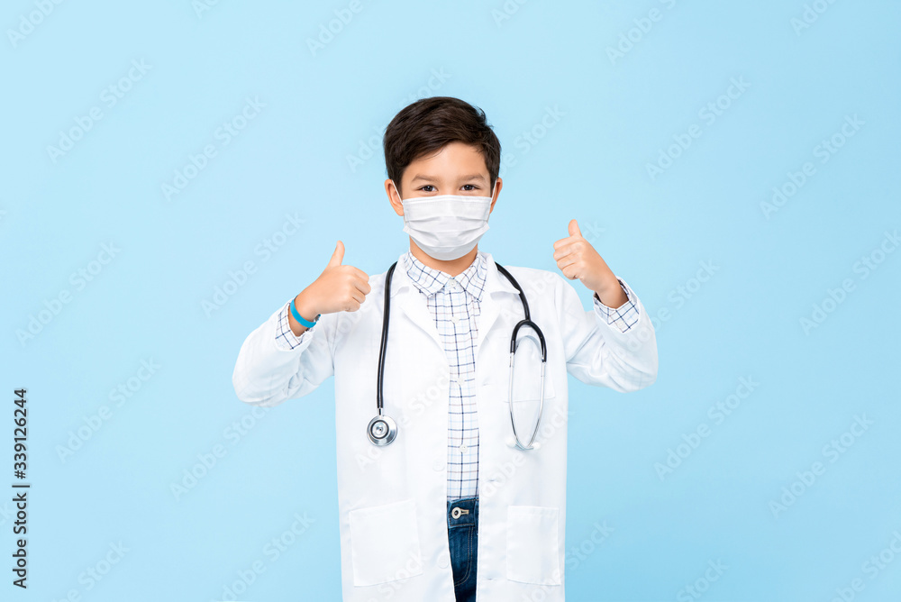Cute boy doctor wearing medical mask and giving thumbs up isolated on light blue background ...