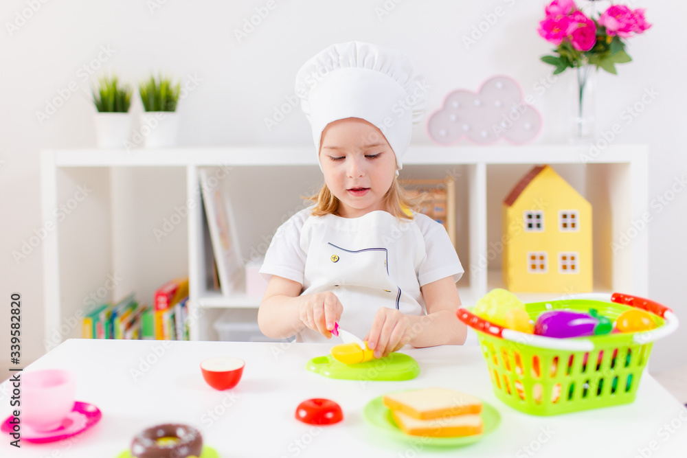 Little blonde girl in white cook uniform playing with toy fruits and ...