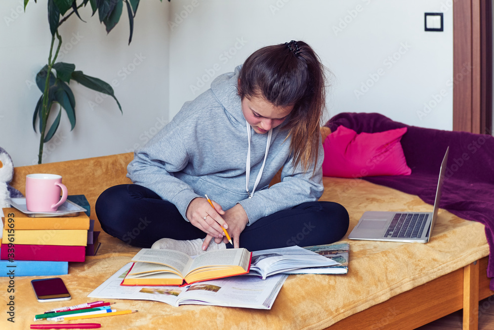 Teenage girl studying at home sitting on bed reading book concentrating ...
