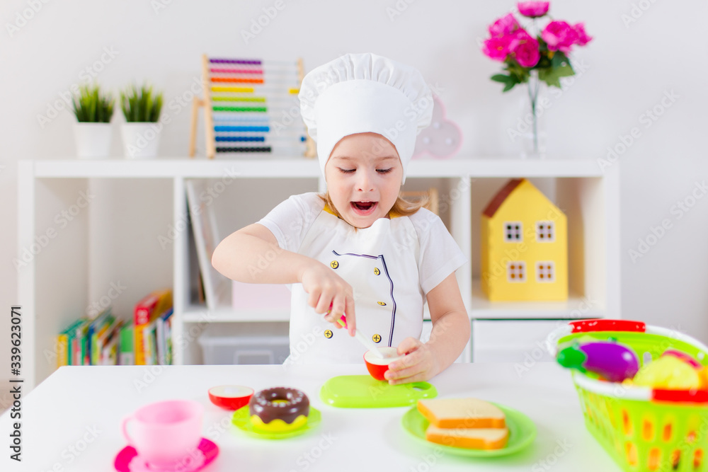 Little blonde girl in white cook uniform playing with toy fruits and ...