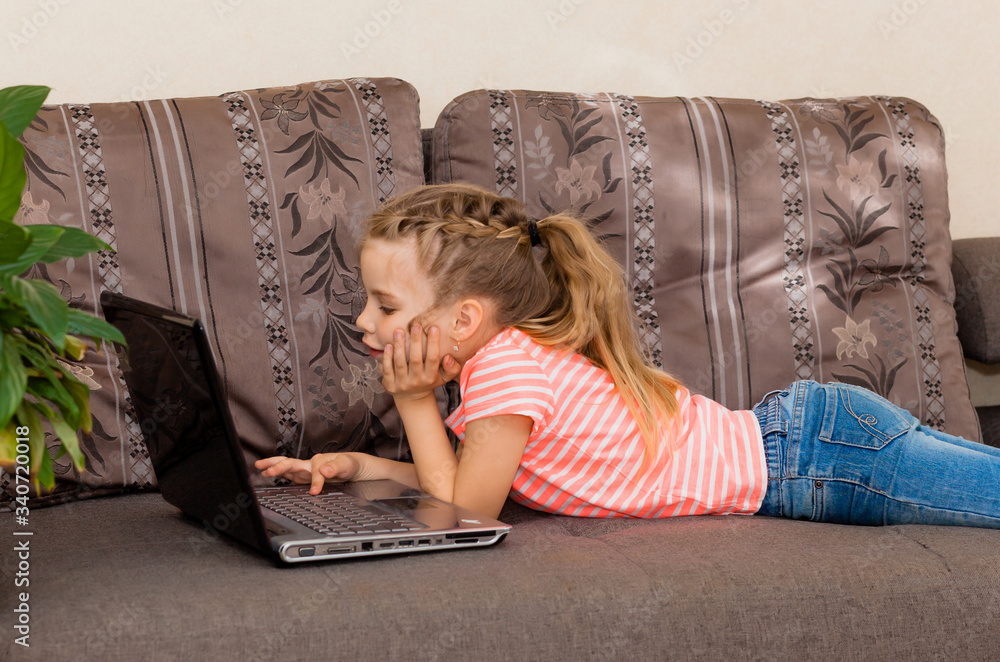 Distance learning. Smiling little girl sitting on a sofa with a laptop ...