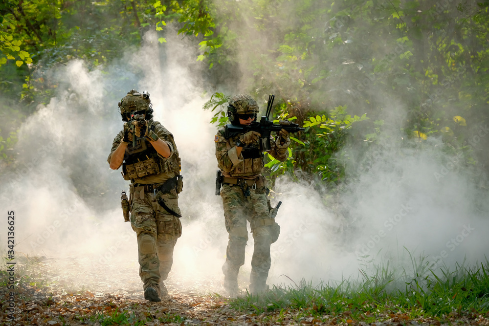 Two soldiers with the fighting uniform walk on the ground and point gun ...