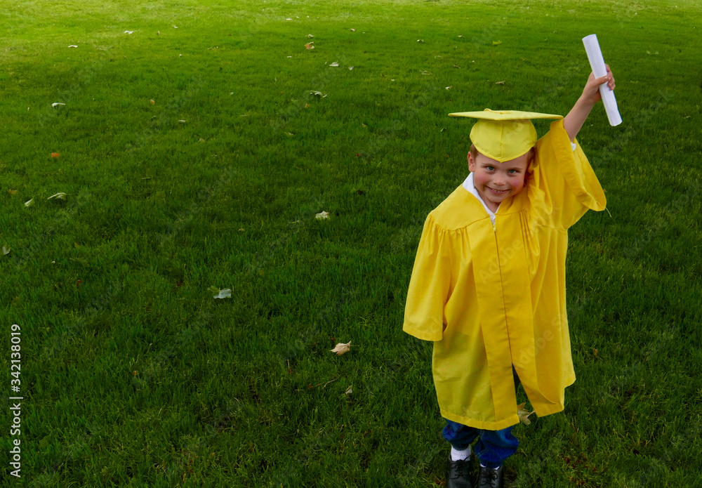 Young boy in a yellow cap and gown smiles and holds up his preschool ...