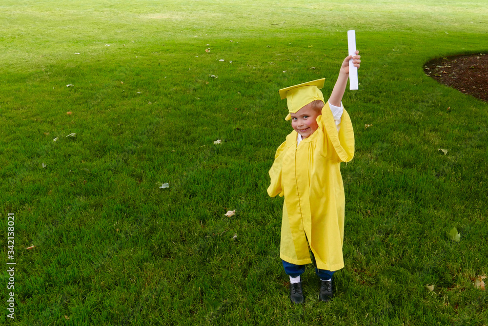 Young boy in a yellow cap and gown smiles and holds up his preschool ...