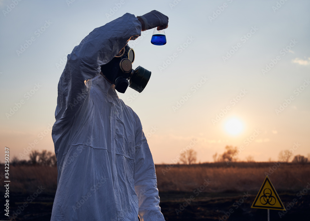 Scientist in white coverall, gas mask and gloves holding test tube with ...