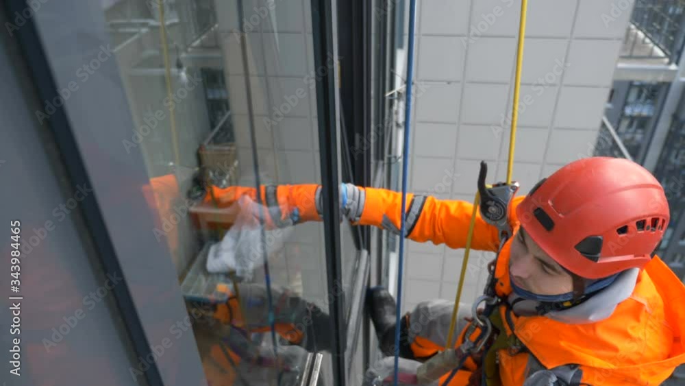 equipped window washer in a helmet and orange jumpsuit washes glass ...