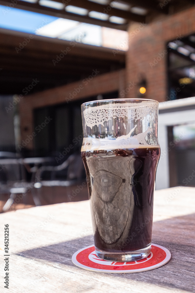 A large clear beer glass of dark ale sits on a table in a micro brewery ...